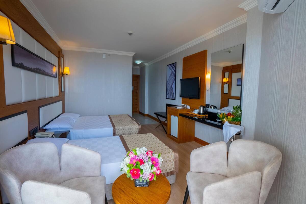 Quarto de hotel com duas camas, uma escrivaninha, uma cadeira, uma mesinha de centro com um vaso de flores e uma televisão.