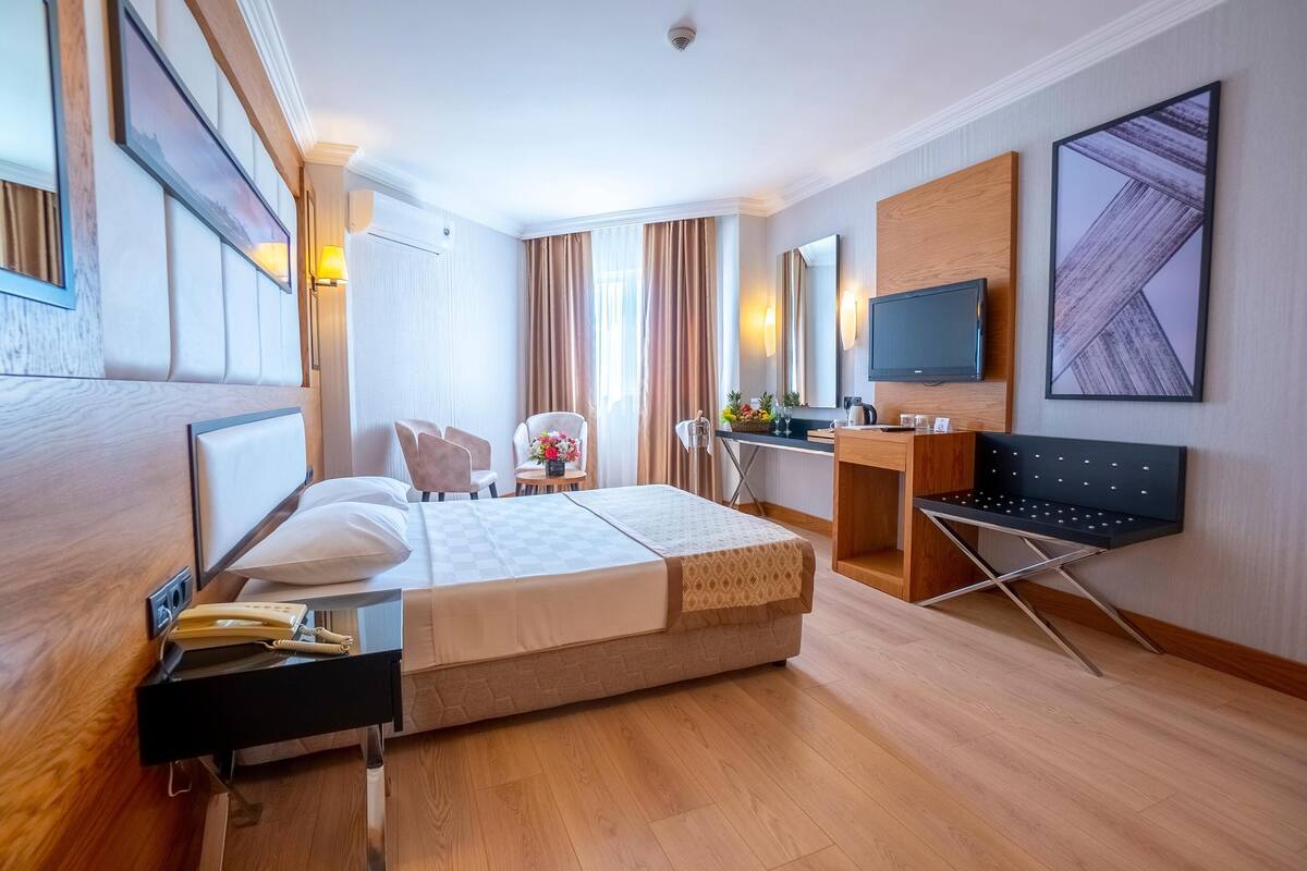 Quarto de hotel com cama, mesa de cabeceira, cadeira, escrivaninha, televisão e obras de arte nas paredes.