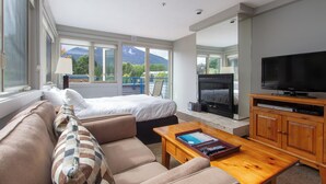 Quarto de hotel moderno com uma cama grande, um sofá, mesa de centro de madeira, televisão e uma varanda com vista para as montanhas.
