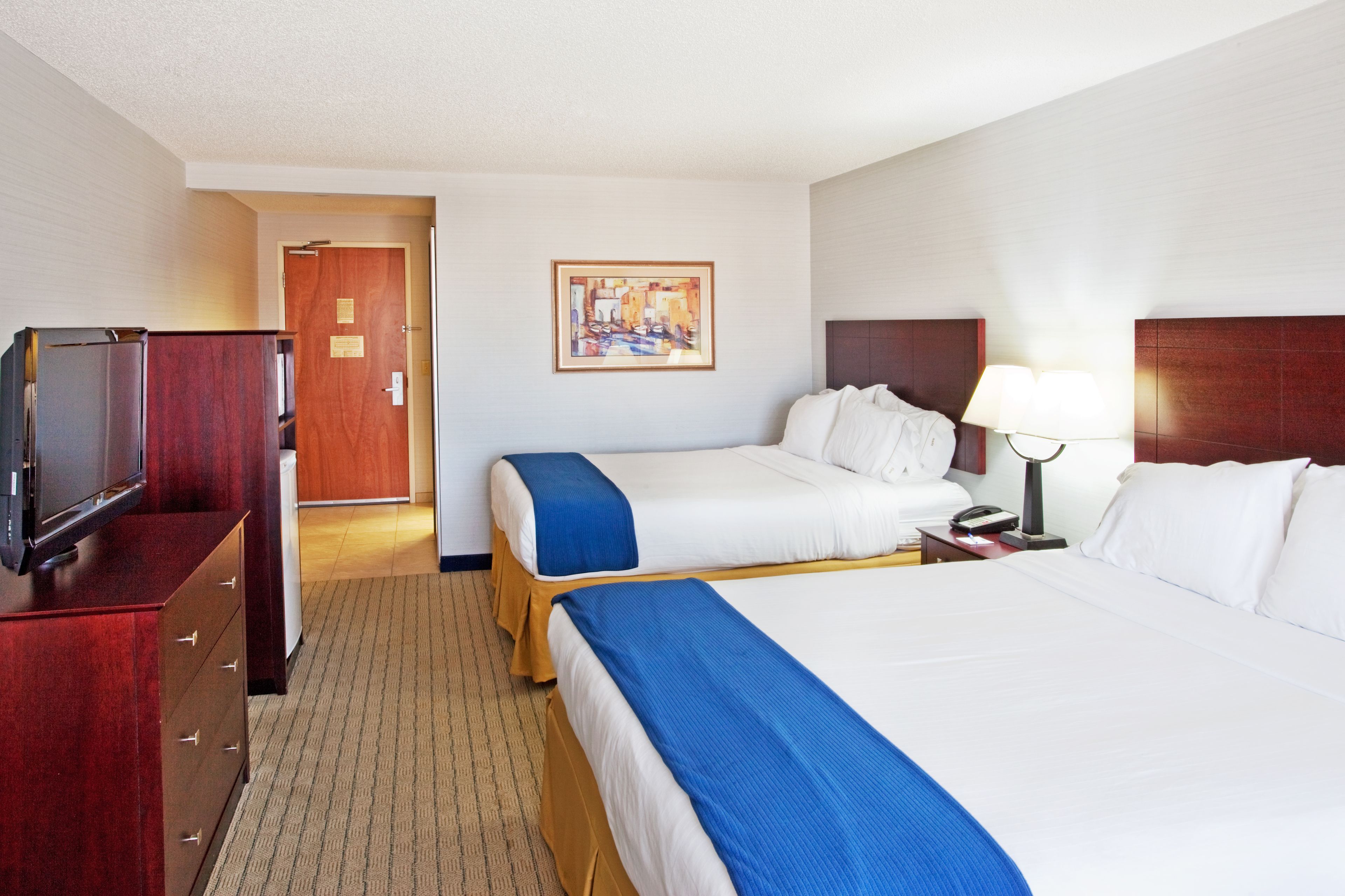 Quarto de hotel com duas camas, televisão, secretária com abajur e um quadro na parede.