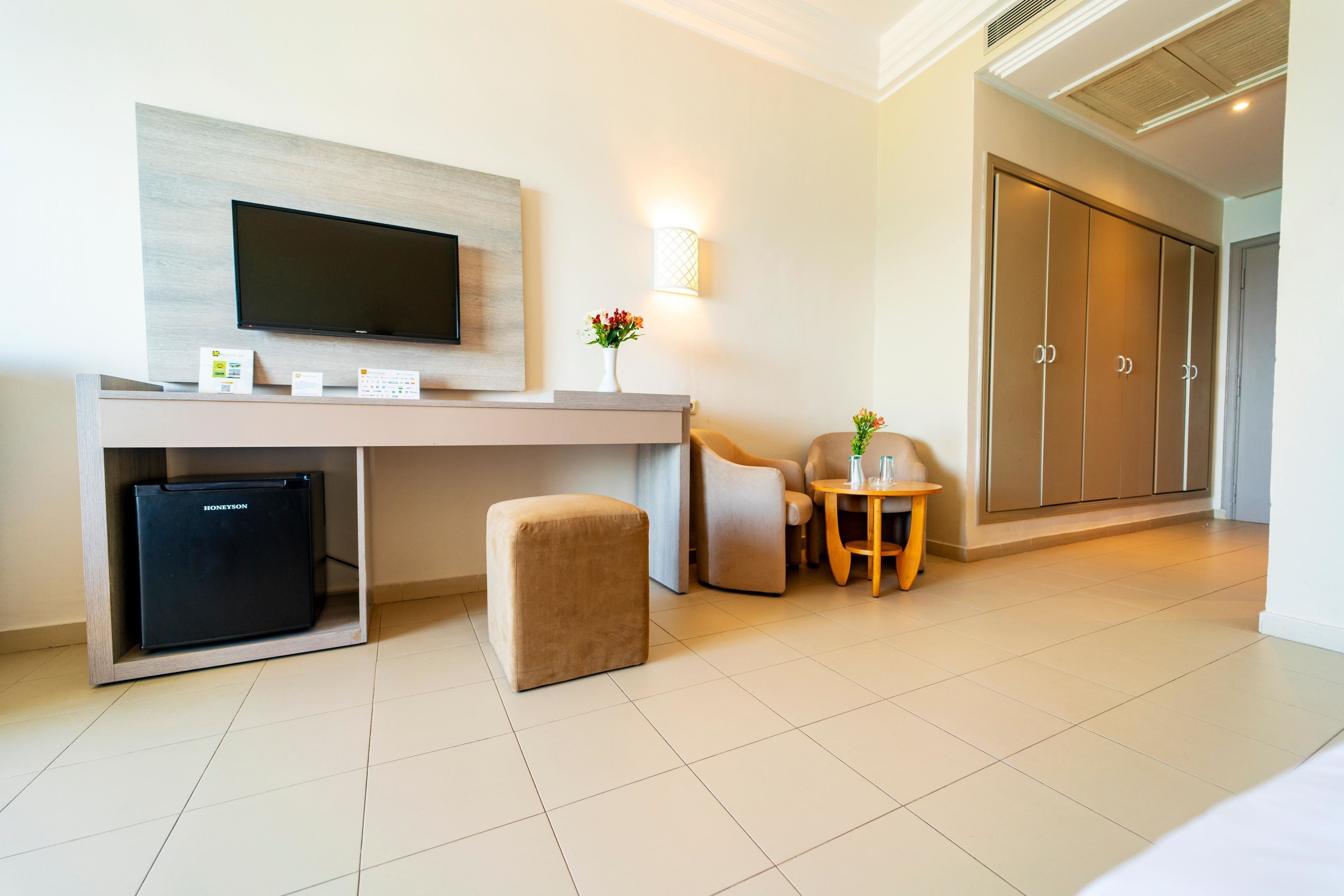 Quarto de hotel moderno com televisão de tela plana, minibar, área de estar com uma pequena mesa e cadeiras, e um guarda-roupa.