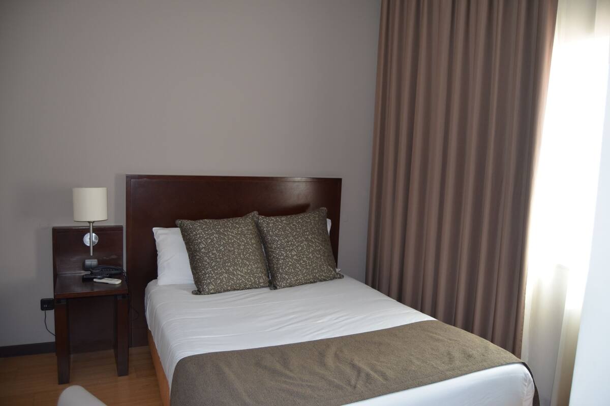 Quarto de hotel com cabeceira de madeira, duas almofadas estampadas, colcha branca, manta marrom, abajur e telefone em uma mesa de cabeceira de madeira.