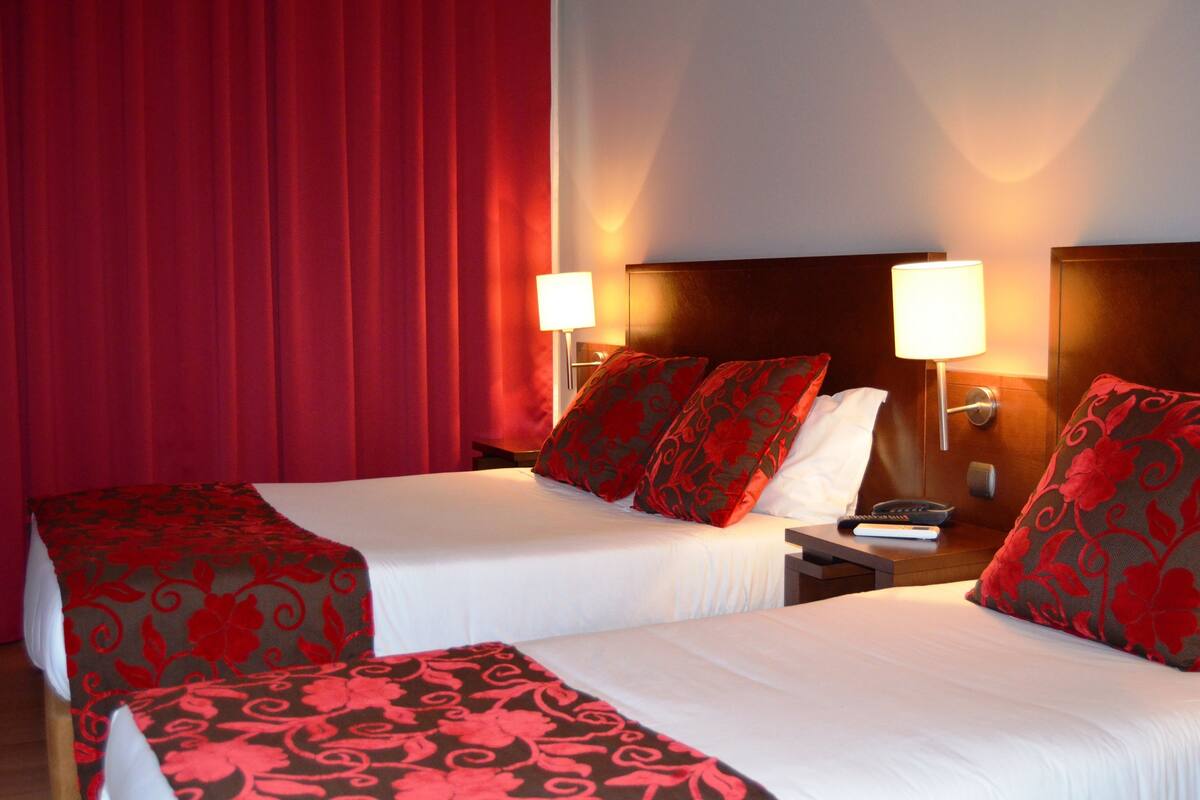 Quarto de hotel com duas camas, cortinas vermelhas e luminárias de cabeceira.