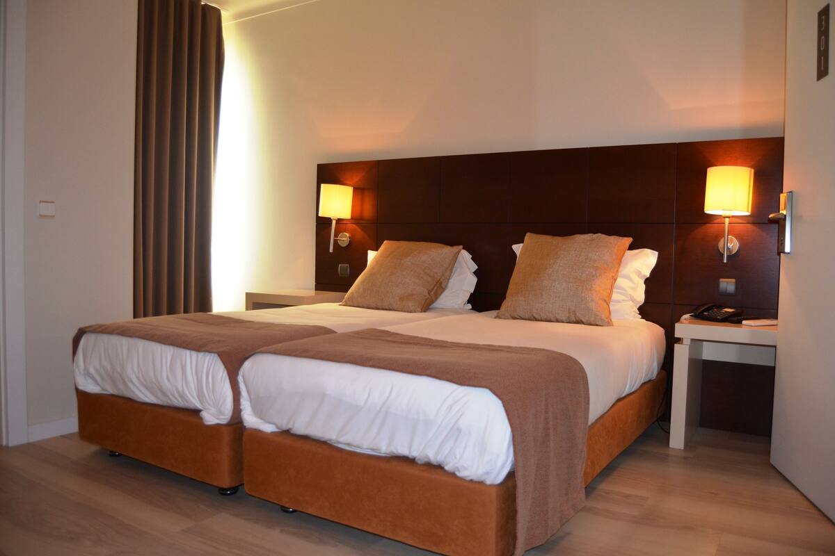 Uma cama de casal com duas almofadas, uma mesa de cabeceira com abajur e uma janela grande com cortinas.