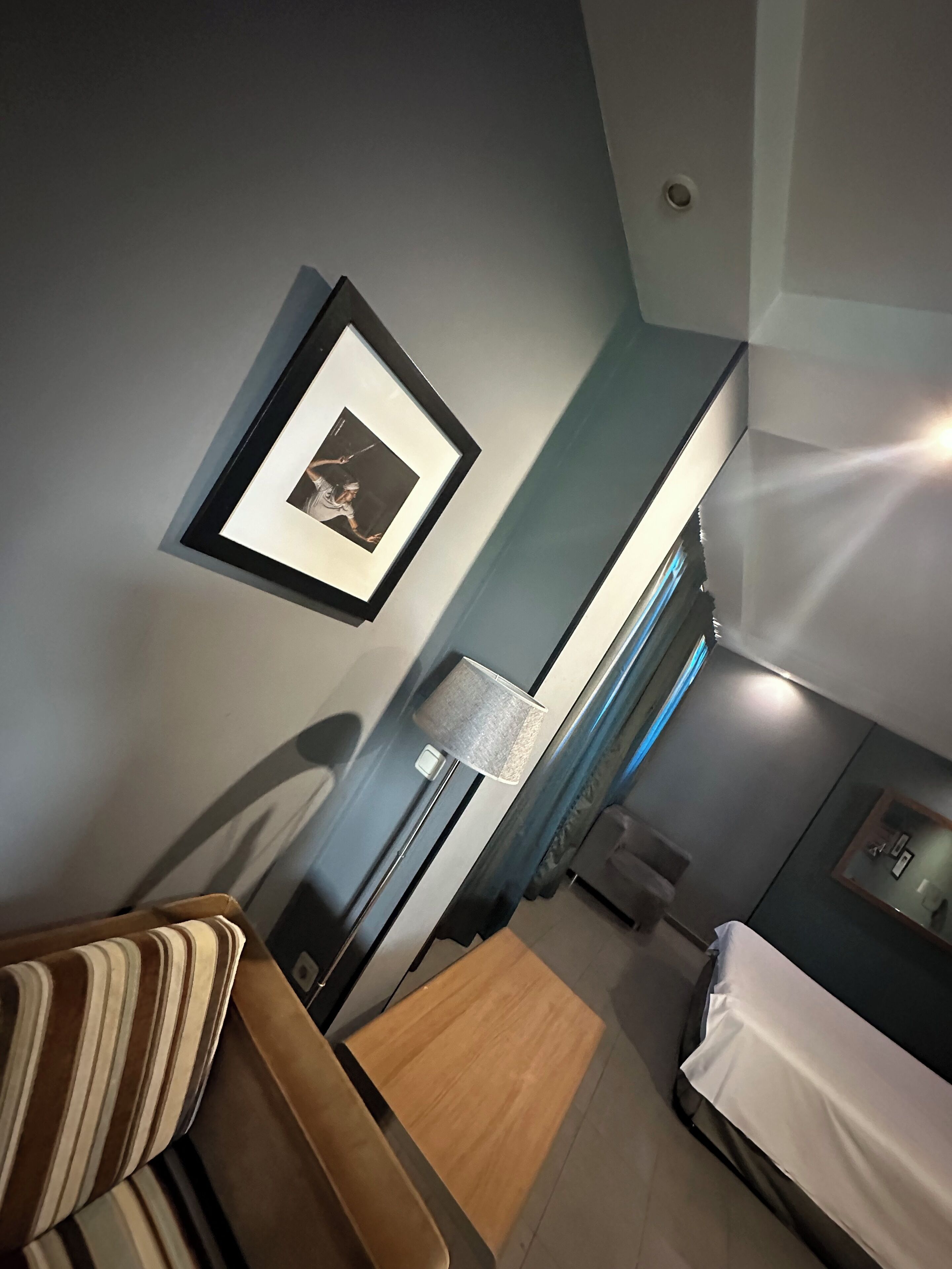 Quarto de hotel moderno com sofá, mesa pequena, quadro na parede, abajur e cama com cobertura branca.