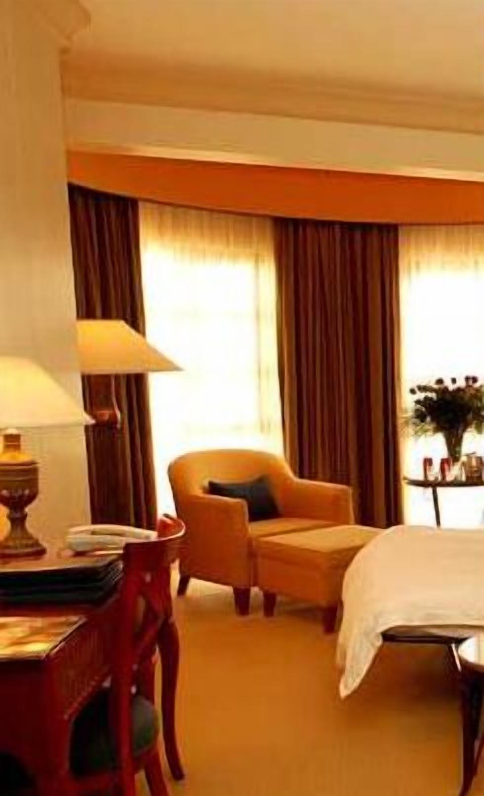 Quarto de hotel com cama, escrivaninha com luminária, cadeira e janela com cortinas.