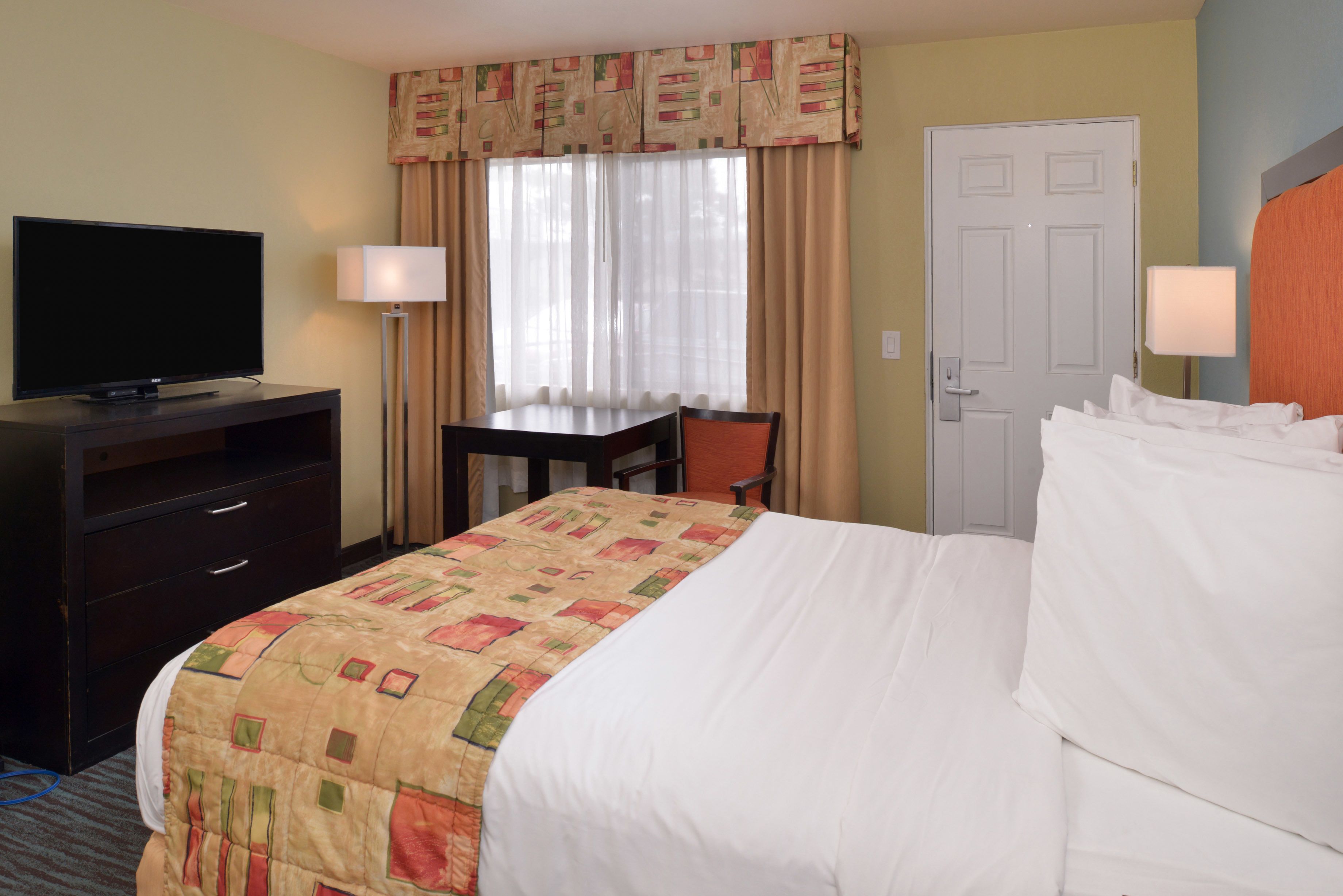 Quarto de hotel com cama, escrivaninha, televisão e porta.
