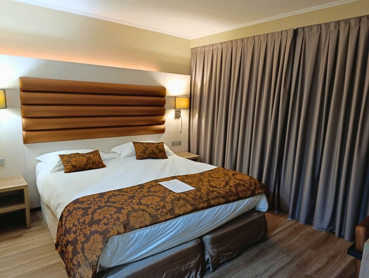 Quarto de hotel com uma cama grande, cabeceira, mesa de cabeceira, abajur e cortina.