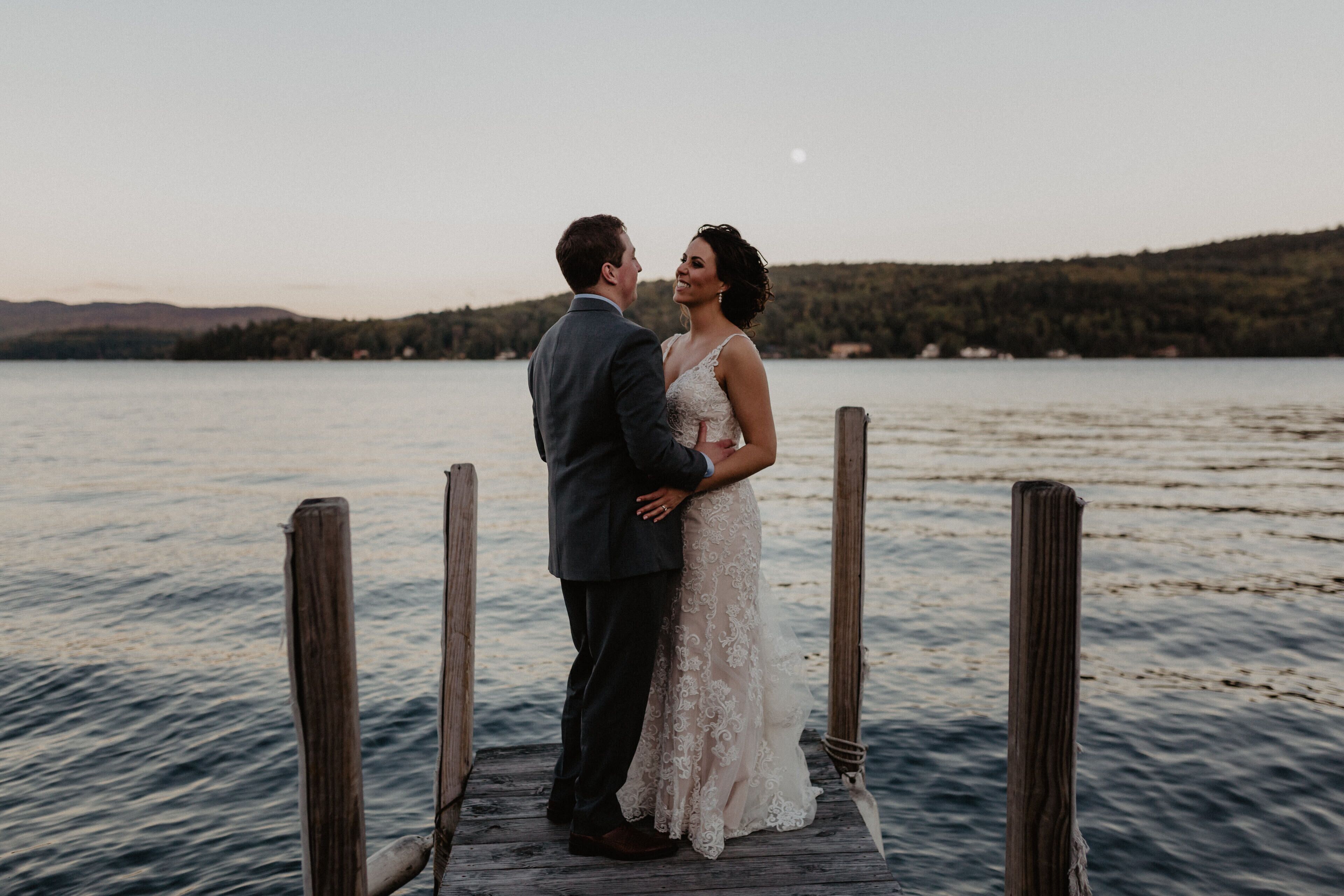 Um casal se abraçando em um cais de madeira à beira de um lago, com colinas ao fundo.