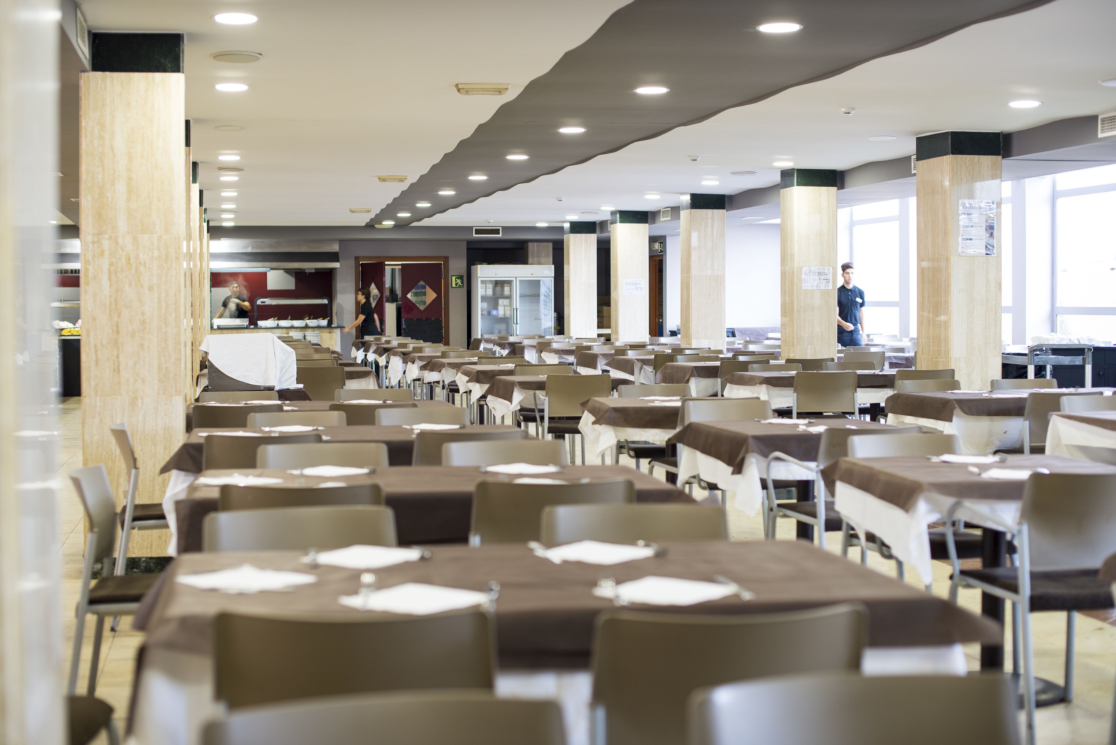 Uma ampla sala de jantar com várias mesas e cadeiras, pilares imponentes e uma pessoa andando ao fundo.