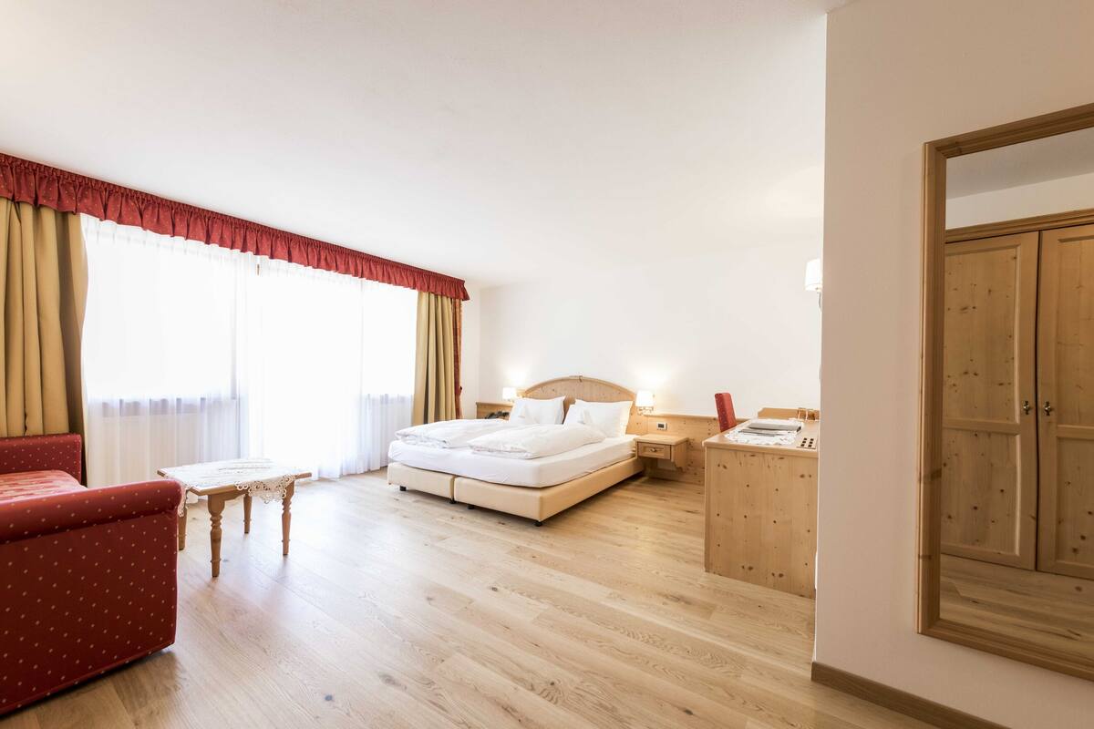 Quarto de hotel com uma cama, um guarda-roupa de madeira, uma mesinha, um sofá vermelho e uma janela com cortinas.