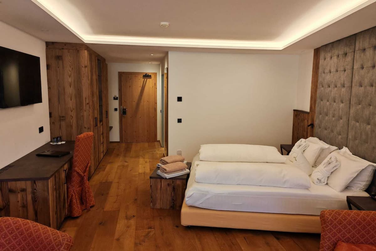 Quarto de hotel moderno com piso de madeira, cama com roupa de cama branca, escrivaninha com cadeira e televisão fixada na parede.