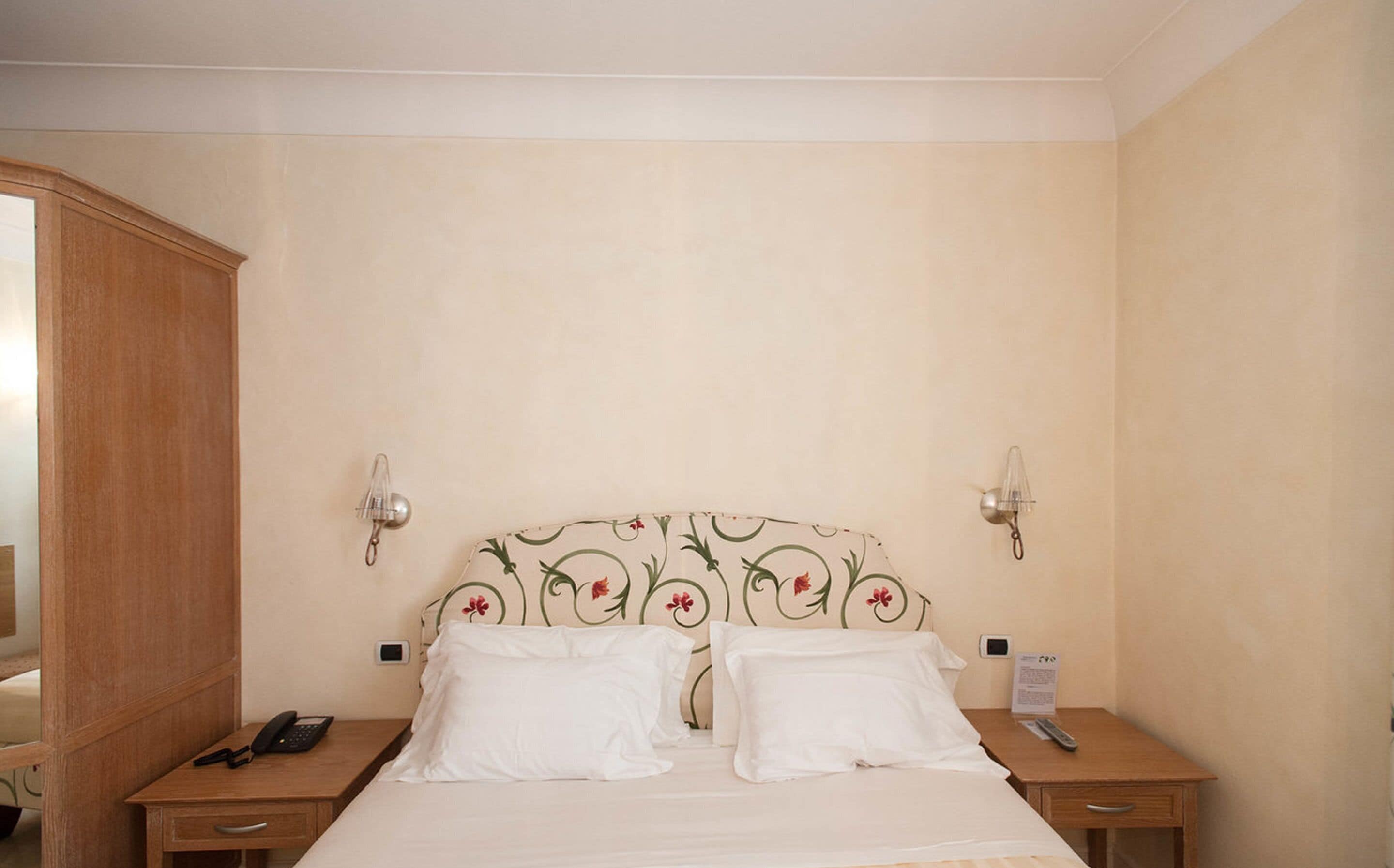 Quarto de hotel com uma cama, duas mesas de cabeceira, um criado-mudo, um espelho e um guarda-roupa de madeira.