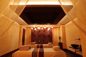 Uma sala de spa com duas camas, uma mesa de massagem fixada na parede, uma pia com cuba e um vaso decorativo com flores.