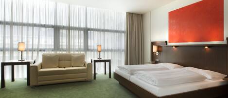 Quarto de hotel com cama, sofá, duas luminárias, uma janela grande com cortinas leves e uma parede em destaque na cor vermelha.