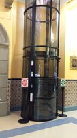 Um elevador cilíndrico com painéis de vidro, situado em um corredor com ladrilhos estampados e um retrato emoldurado na parede.