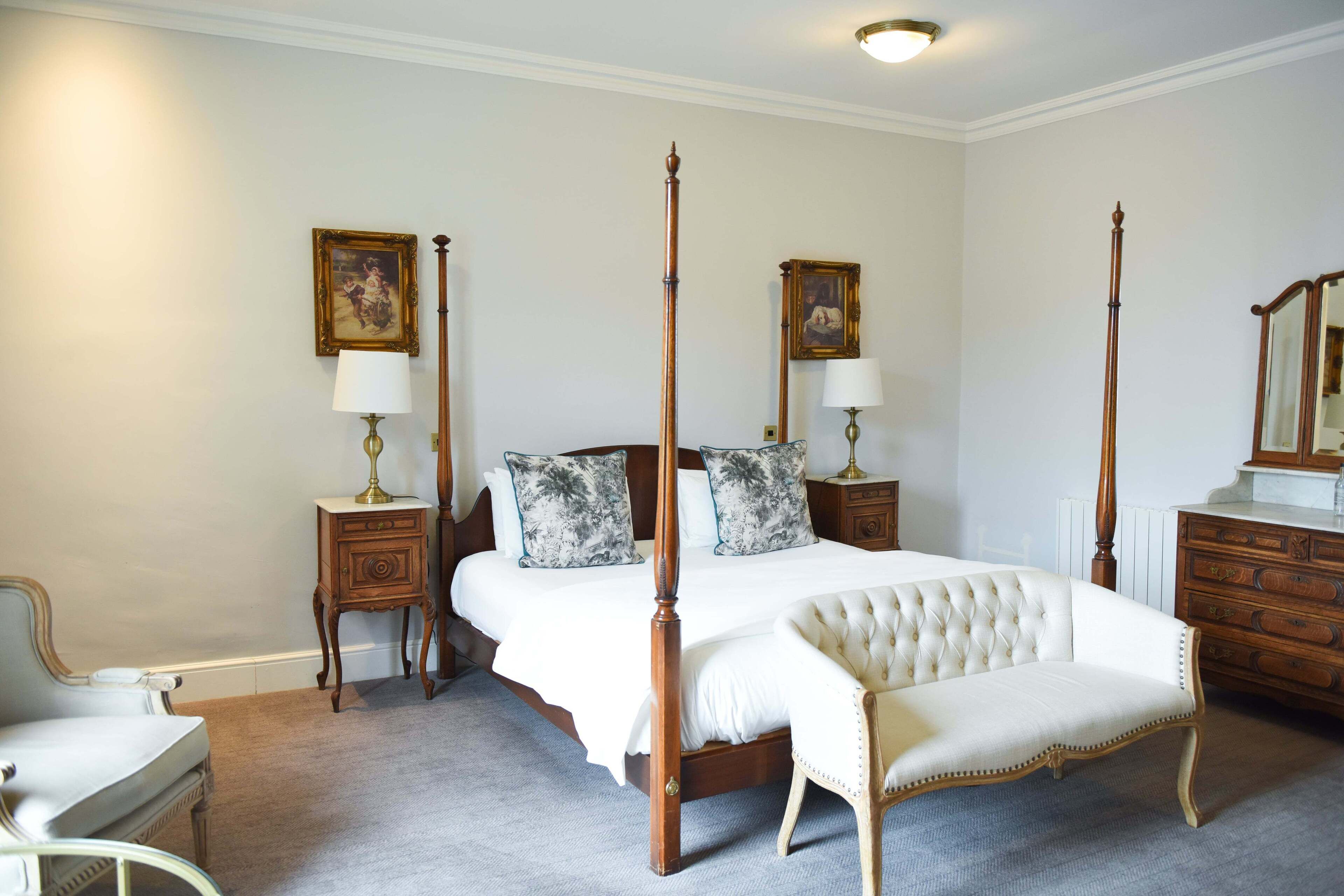 Uma cama de dossel com cabeceira estofada em branco, acompanhada por duas mesas de cabeceira com abajures, um aparador de madeira e uma cadeira.