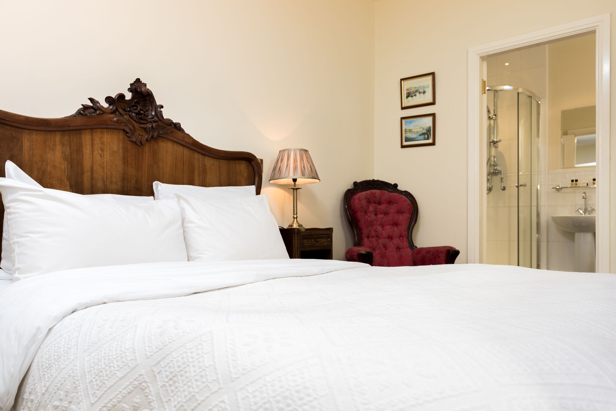 Cama bem arrumada com lençóis brancos, cabeceira de madeira, poltrona vermelha, abajur e porta que dá para um banheiro.