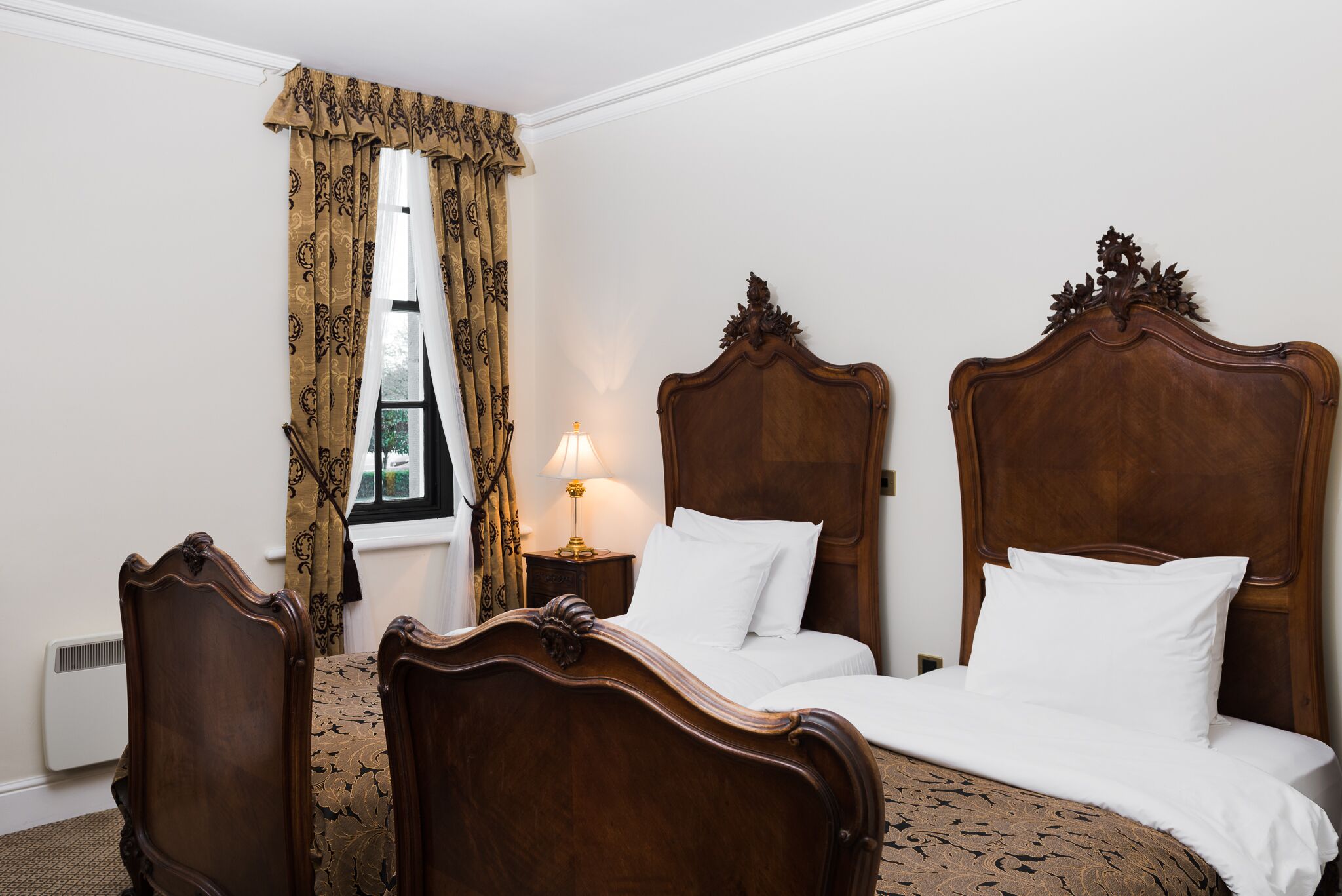 Quarto com cama grande, duas cadeiras, mesa de cabeceira, abajur e janela com cortinas.