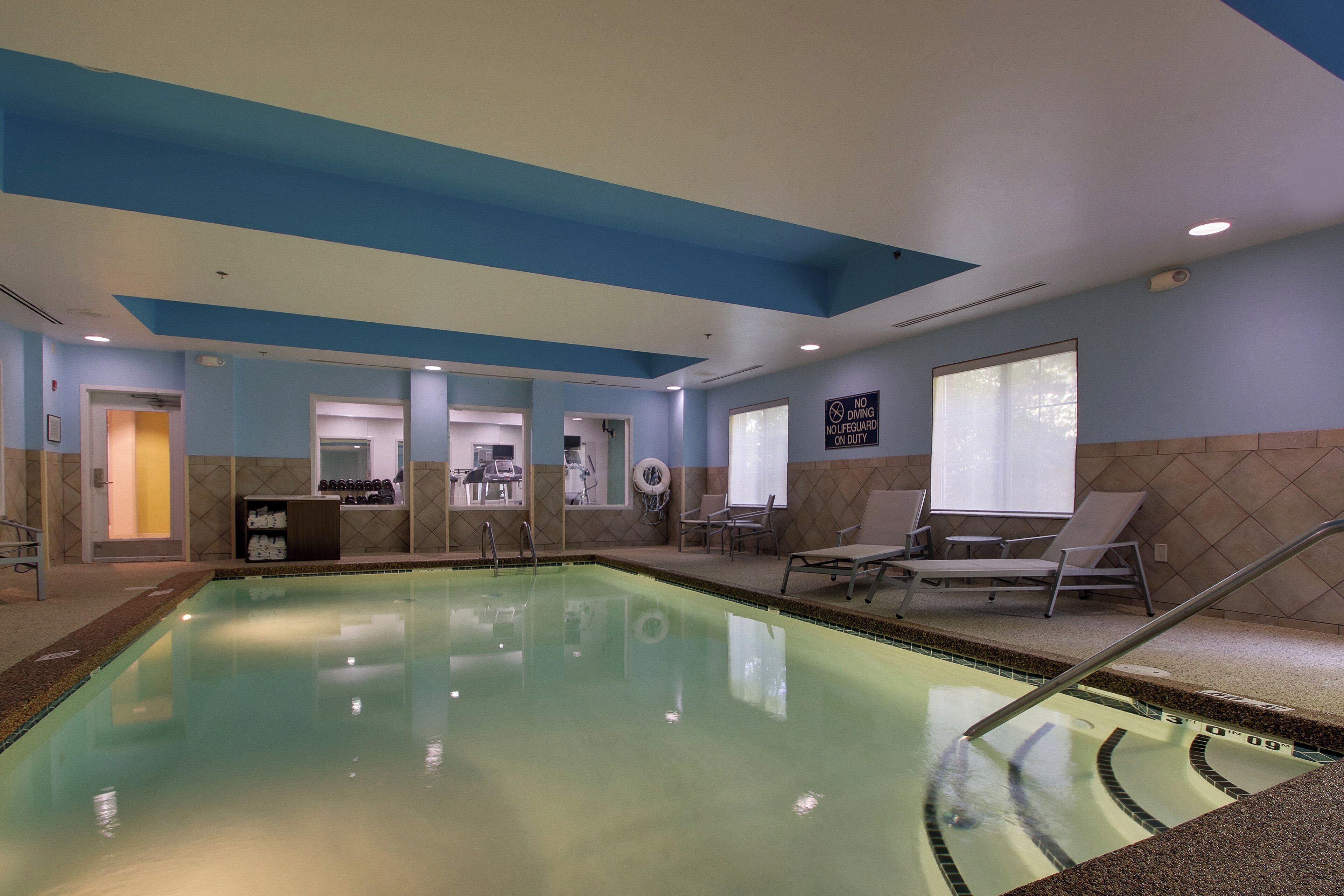 Piscina interna com superfície de água cristalina, placa de salva-vidas e área de estar com cadeiras e mesa.