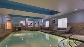 Piscina interna com superfície de água cristalina, placa de salva-vidas e área de estar com cadeiras e mesa.