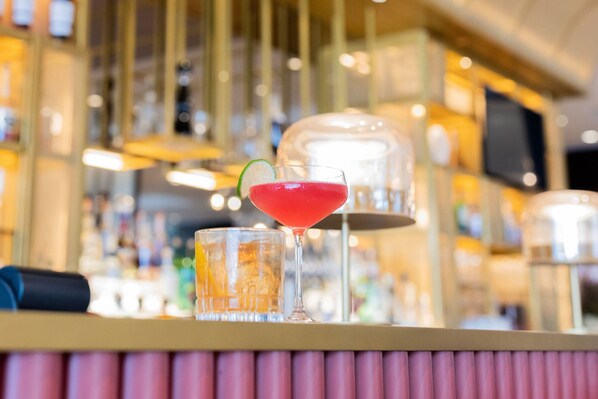 Uma área de bar com um copo de cocktail vermelho e um copo de sumo de laranja.