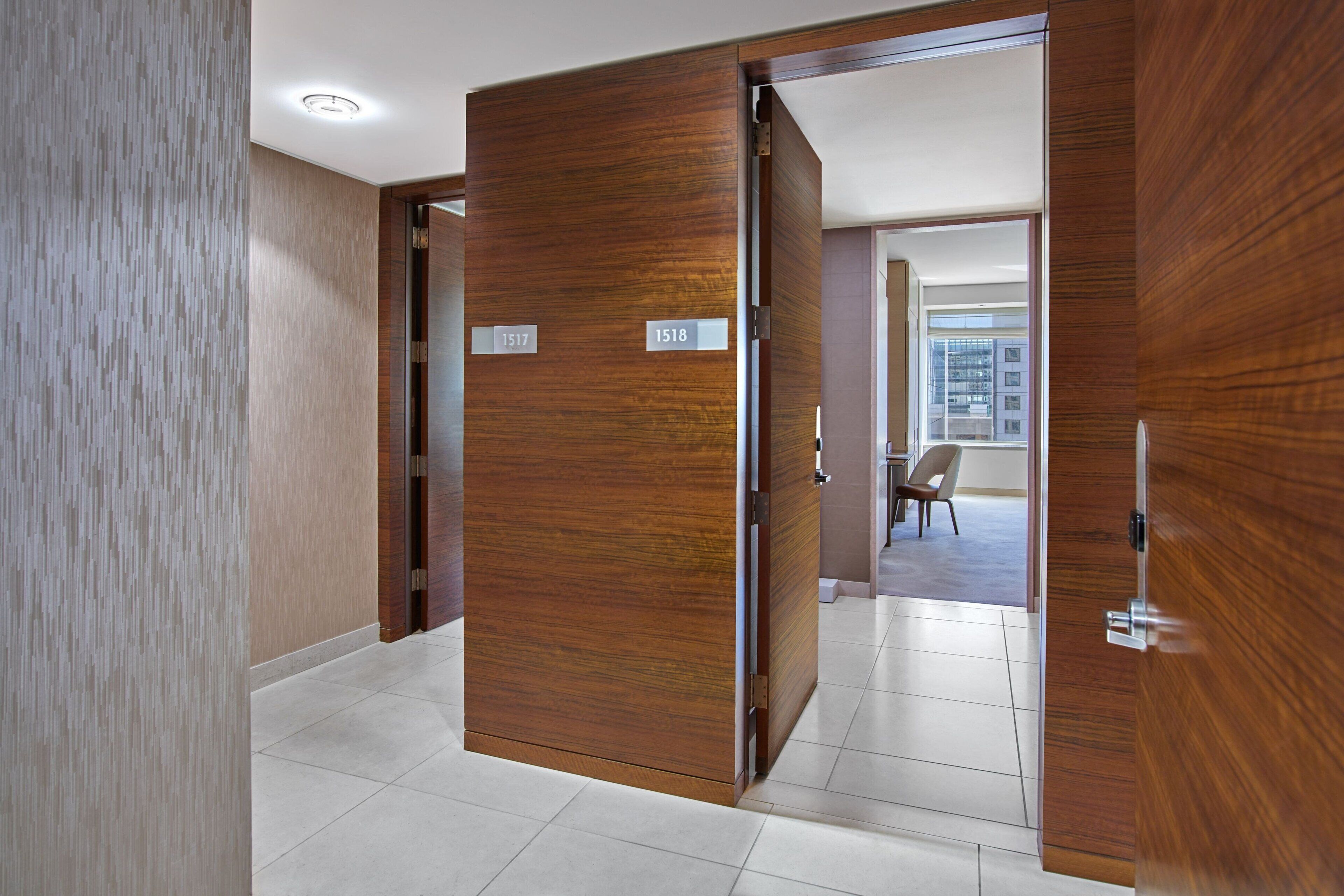 Corredor moderno de hotel, com portas de madeira, placas de identificação de quartos numeradas e vista para uma área de refeições.