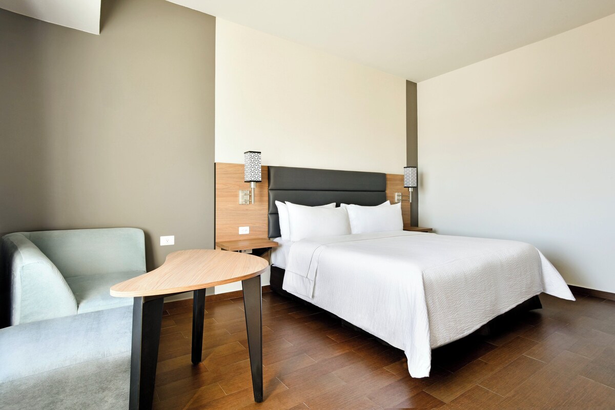 Quarto de hotel com cama, uma cadeira, uma mesinha e mesa de cabeceira com abajures.