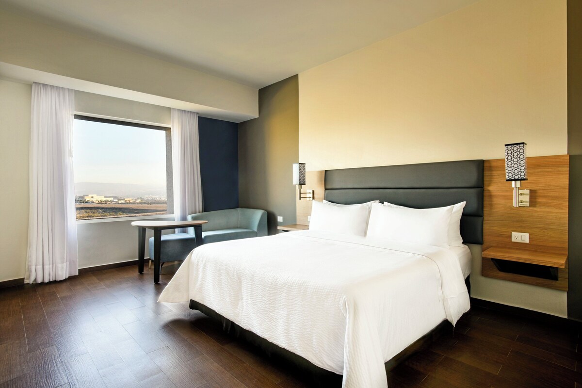 Quarto de hotel com uma cama grande, uma mesa pequena, uma cadeira e vista para a paisagem pela janela.