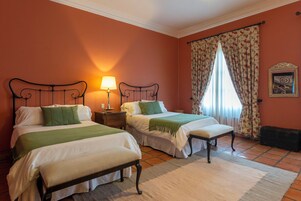 Quarto com duas camas, mesa de cabeceira com abajur, um banco, janela com cortinas florais e um quadro na parede.
