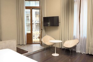 Quarto de hotel com varanda, televisão na parede e duas cadeiras com uma pequena mesa.