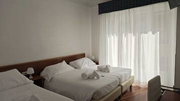 Quarto de hotel com duas camas, painel de cabeceira de madeira, escrivaninha com cadeira e uma janela grande com cortinas leves.