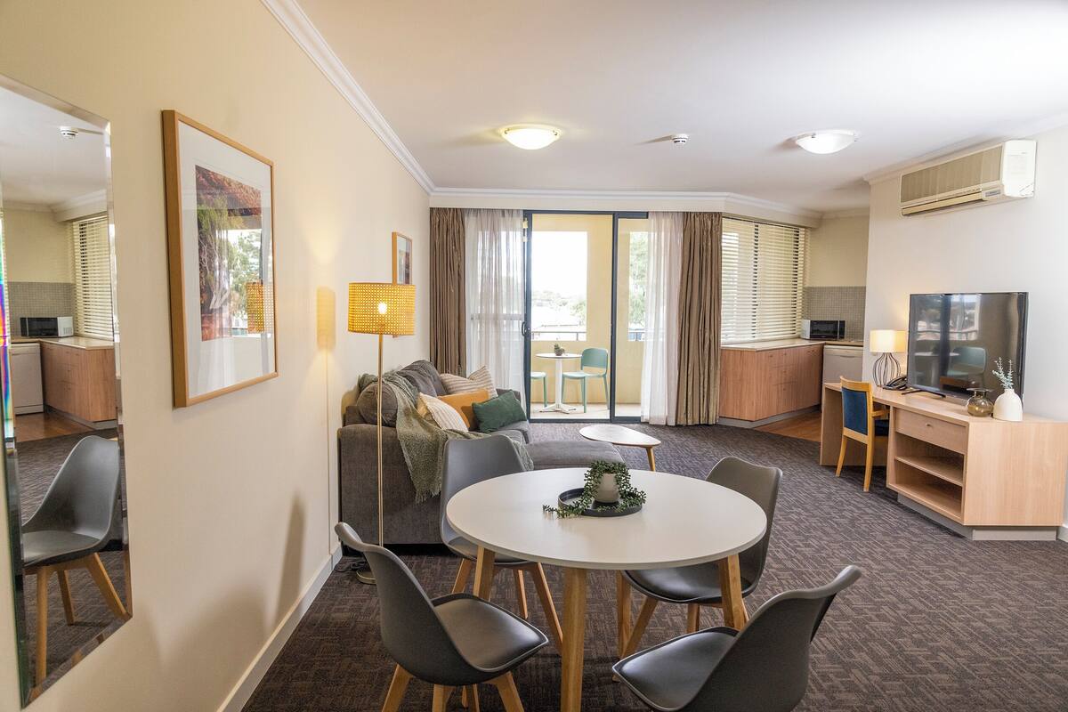 Quarto de hotel moderno com área de refeições, sofá, televisão e cozinha compacta.