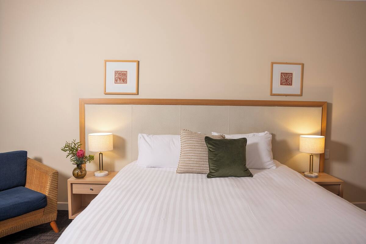 Quarto de hotel com uma cama grande, duas mesas de cabeceira com abajures, uma cadeira de vime e duas imagens emolduradas na parede.