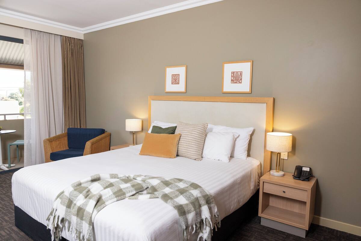 Quarto de hotel com uma cama grande, duas almofadas, uma mesa de cabeceira com abajur e telefone, uma cadeira e duas fotografias emolduradas na parede.