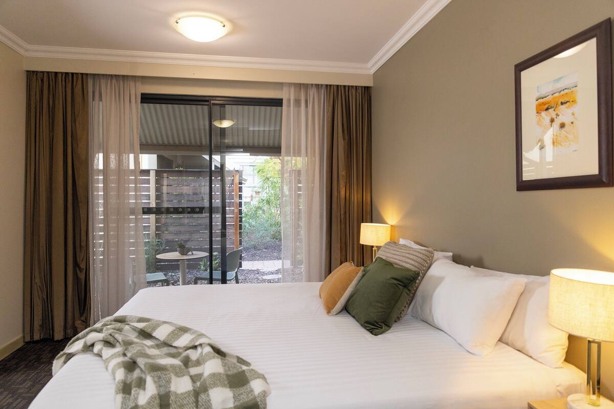 Quarto de hotel com uma cama grande, vista para o exterior por uma porta de correr e uma pequena mesa com uma cadeira do lado de fora.