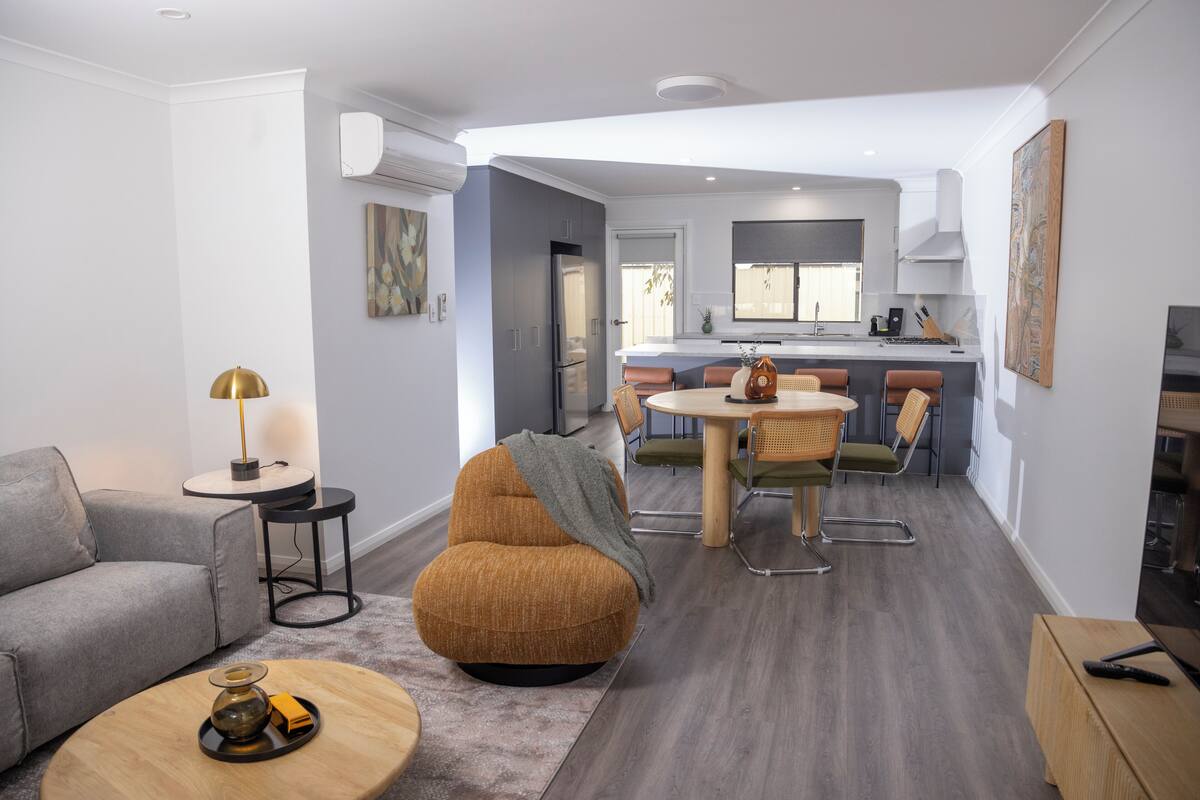 Uma sala de estar moderna com um sofá cinza, uma mesa de centro redonda e uma poltrona laranja única. É visível, ao fundo, uma área de jantar com mesa e cadeiras redondas.