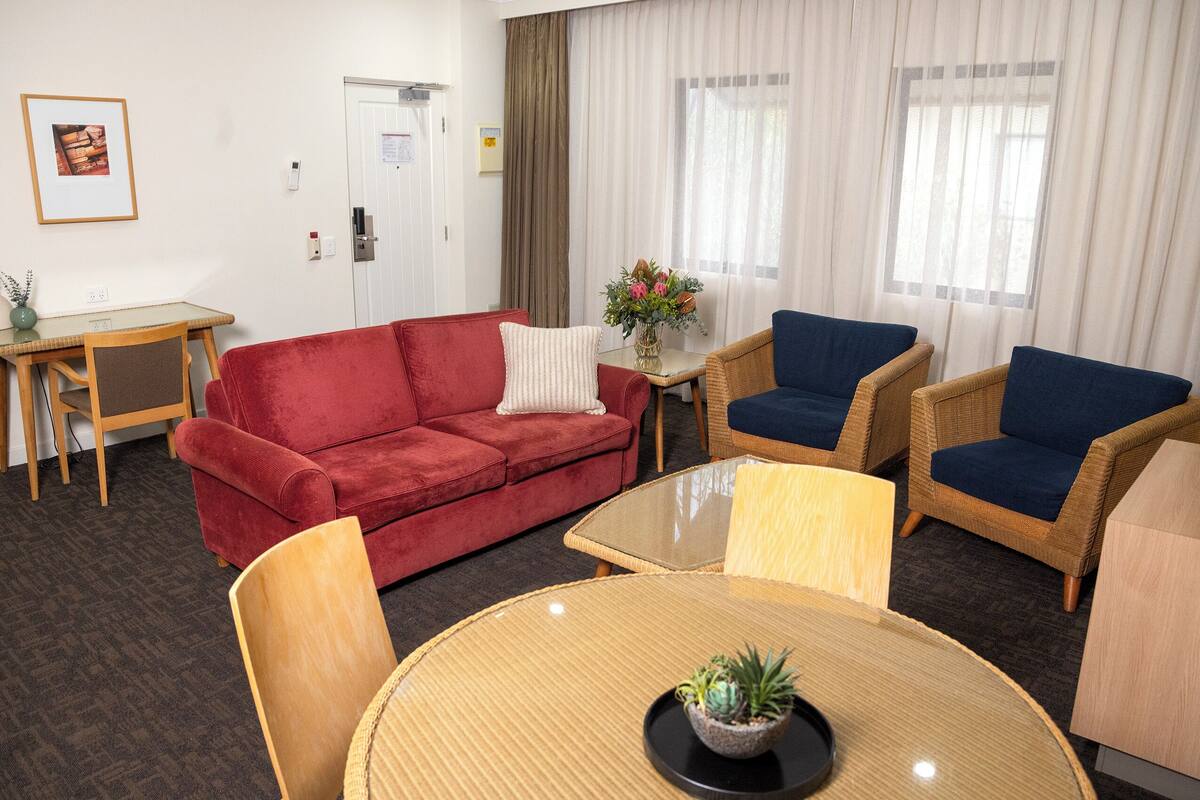 Uma sala de estar com um sofá vermelho, poltronas azuis, uma mesa de centro de madeira com uma planta e uma pequena mesa lateral com um vaso de flores.