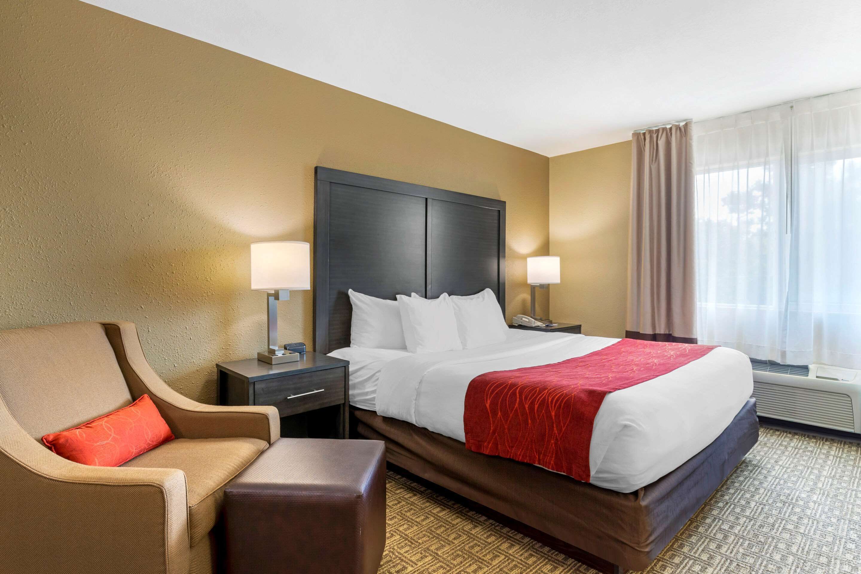 Quarto de hotel com uma cama grande, um sofá, uma mesinha de cabeceira e uma janela com cortinas.