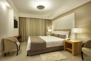 Quarto de hotel moderno com uma cama grande, duas mesas de cabeceira com luminárias, uma cadeira, um sofá e uma janela ampla com cortinas.