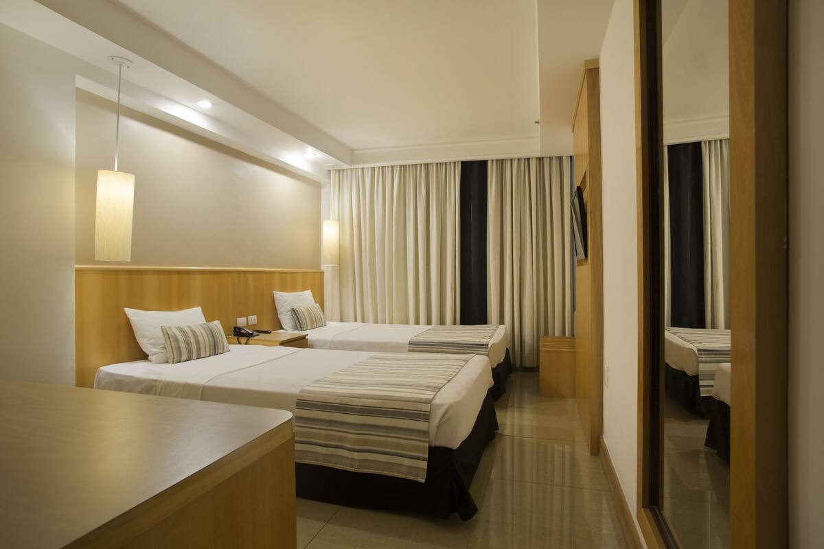 Quarto de hotel com duas camas, cabeceira de madeira, mesa de cabeceira, luminária e um espelho grande.