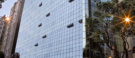 Um edifício alto com fachada de vidro, entrada no nível da rua e uma rua visível com pedestres.