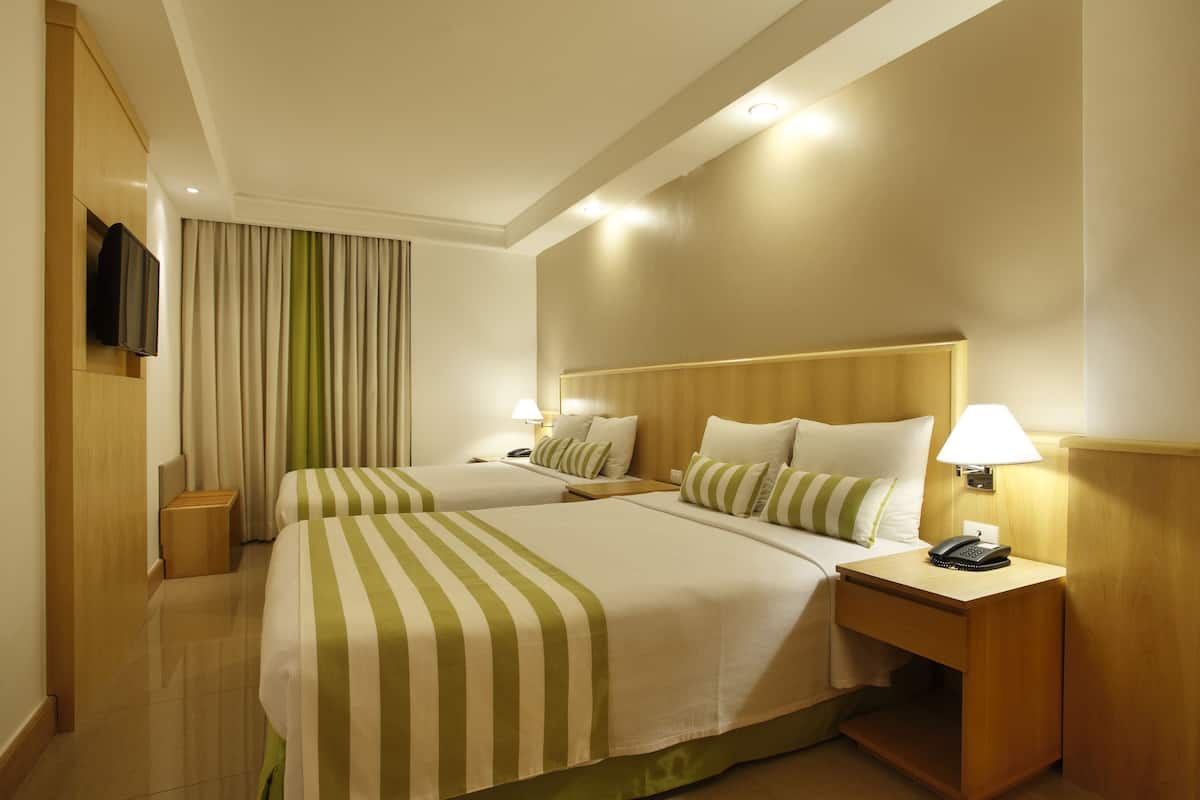 Quarto de hotel com duas camas, cada uma com roupa de cama listrada, mesas de cabeceira com abajures e uma televisão fixada na parede.