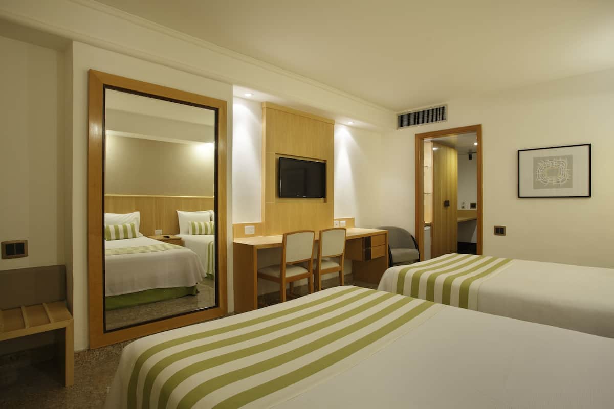 Quarto de hotel com duas camas, uma escrivaninha, uma cadeira, televisão e espelho.
