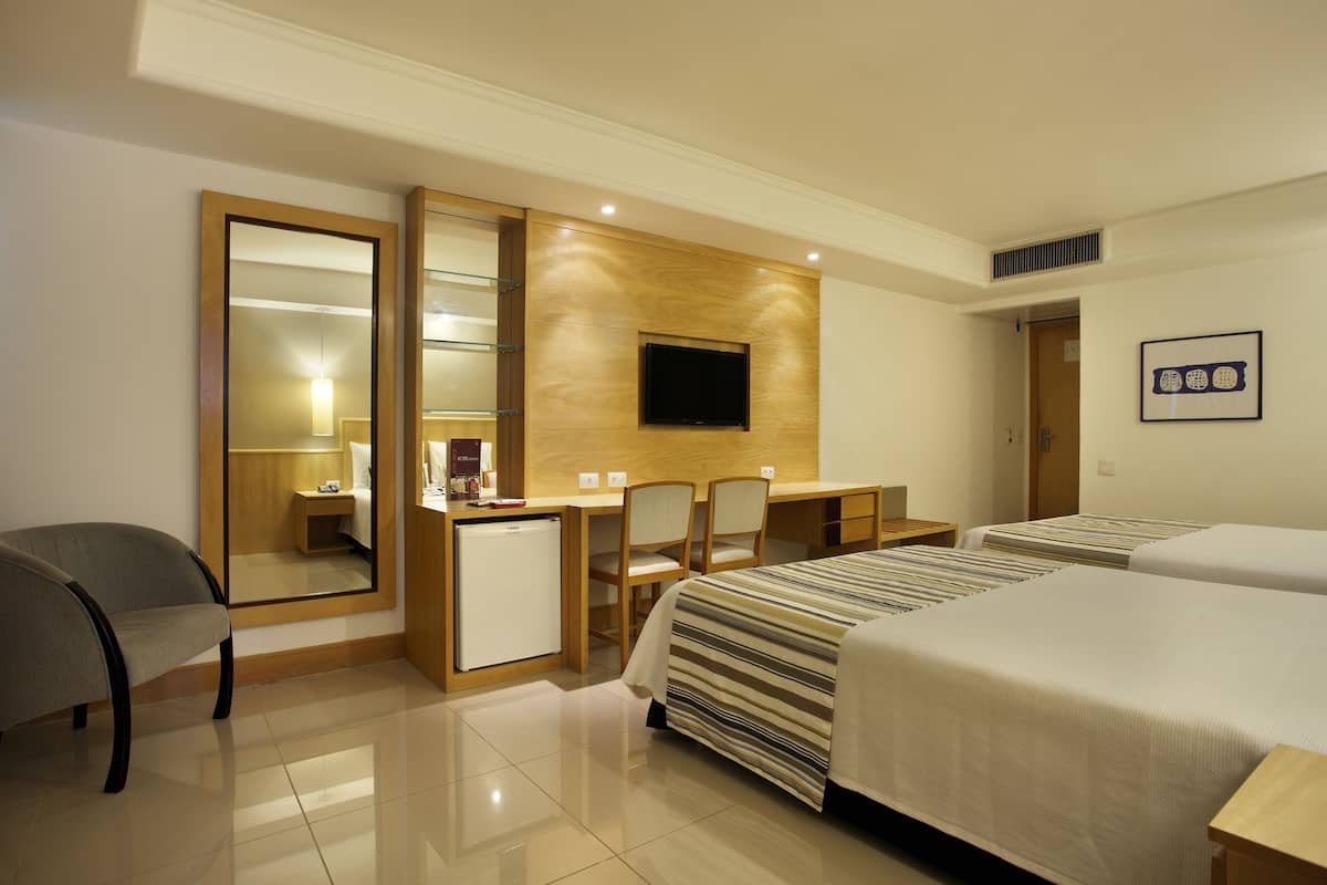 Quarto de hotel com cama, escrivaninha, cadeira, um pequeno frigorífico e televisão na parede.