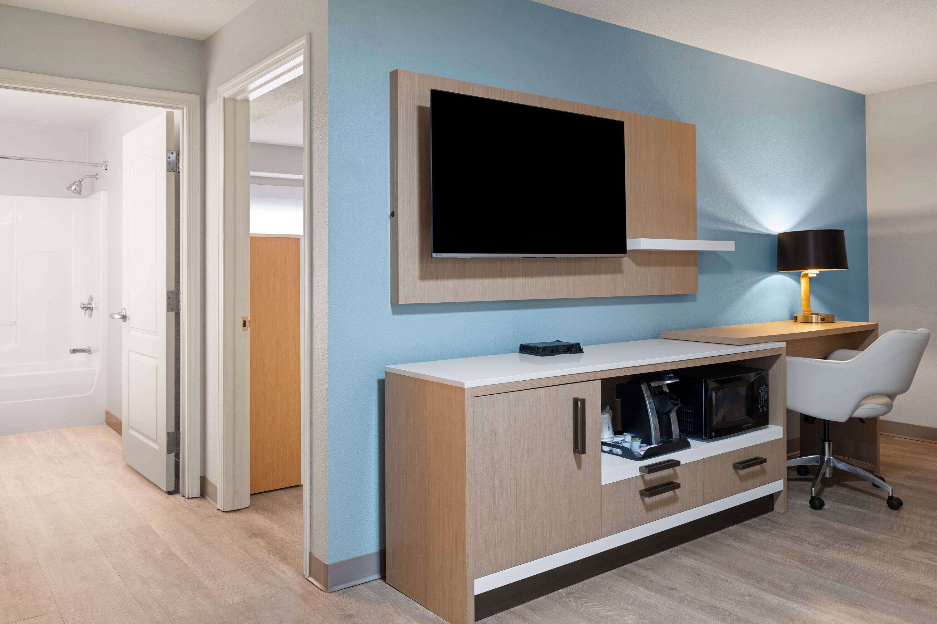 Quarto de hotel moderno com TV de tela plana fixada acima de um armário de madeira, uma escrivaninha com luminária e uma cadeira de escritório branca.