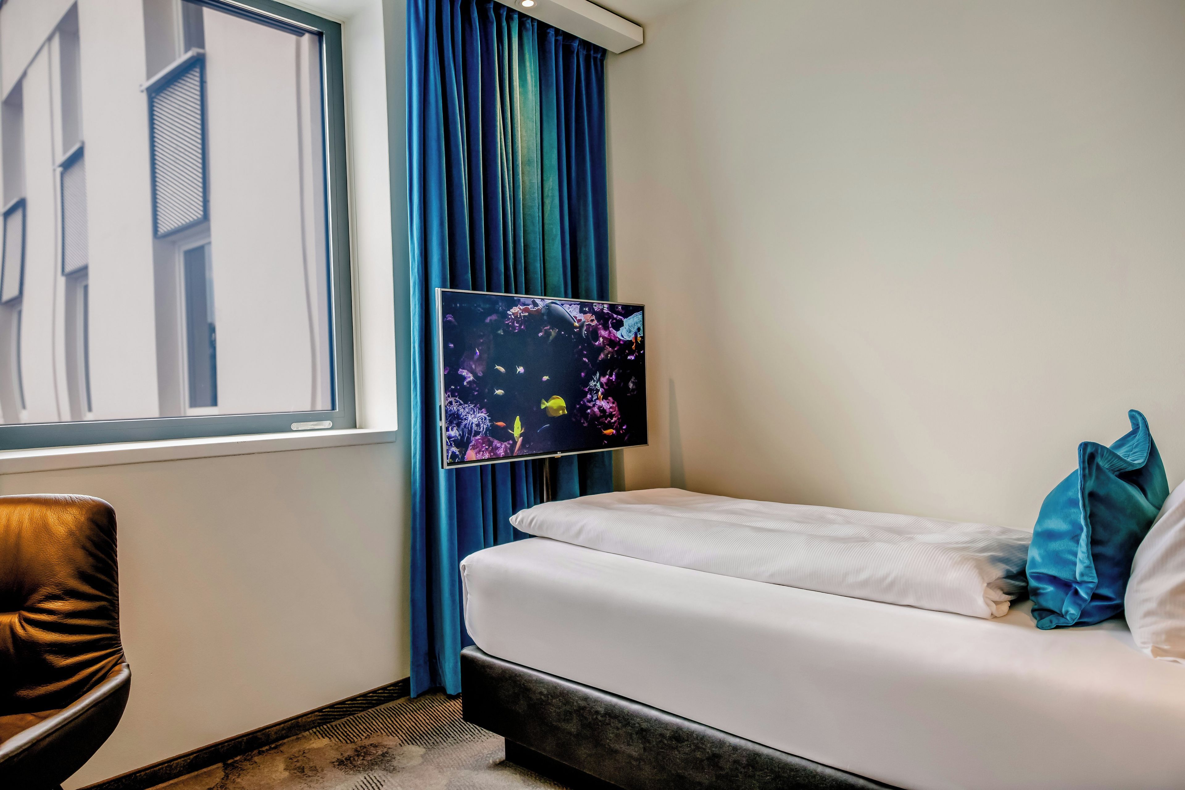 Quarto de hotel com cama, televisão fixada na parede, janela com cortinas e uma cadeira.