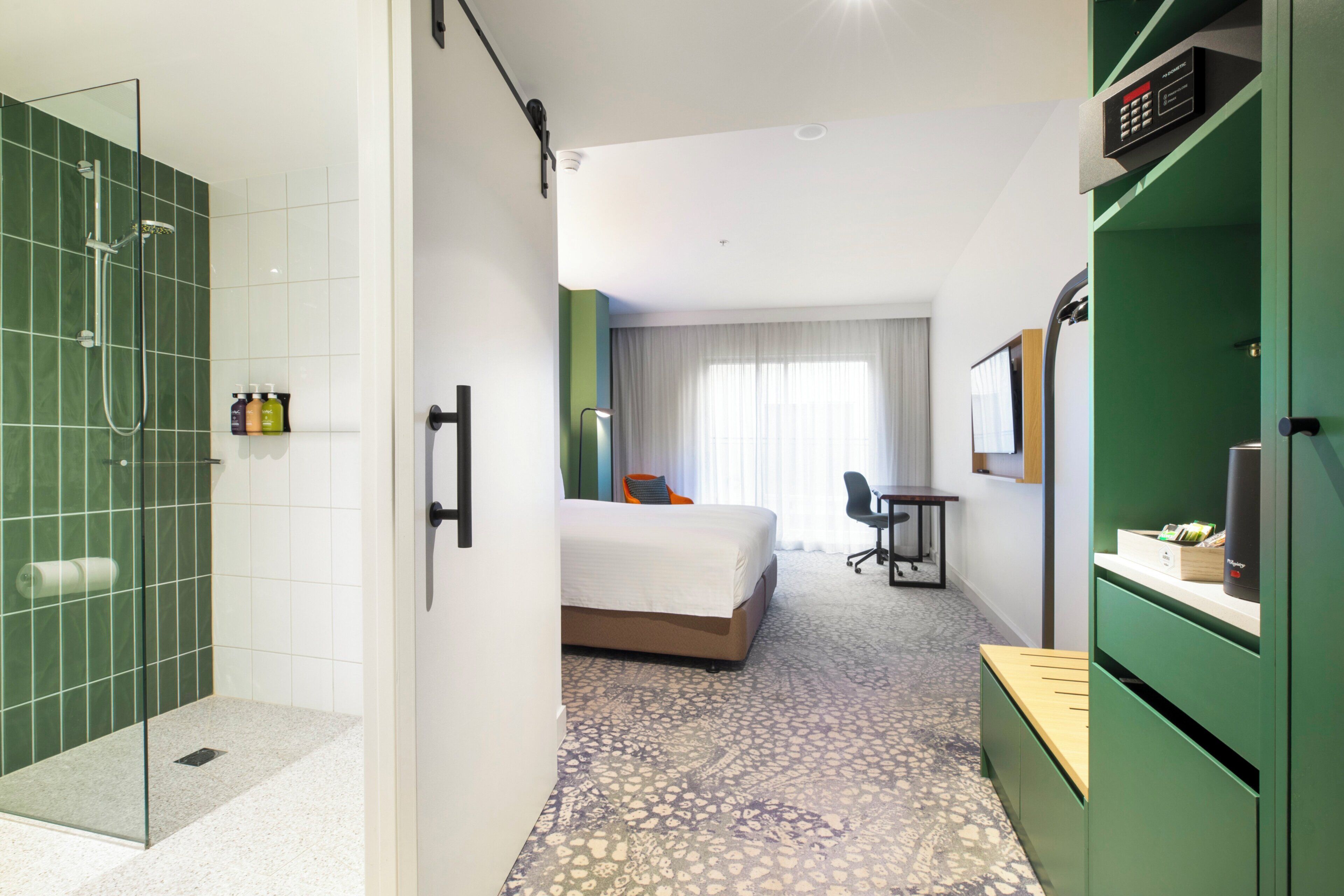 Quarto de hotel com chuveiro, cama, escrivaninha e armários verdes.