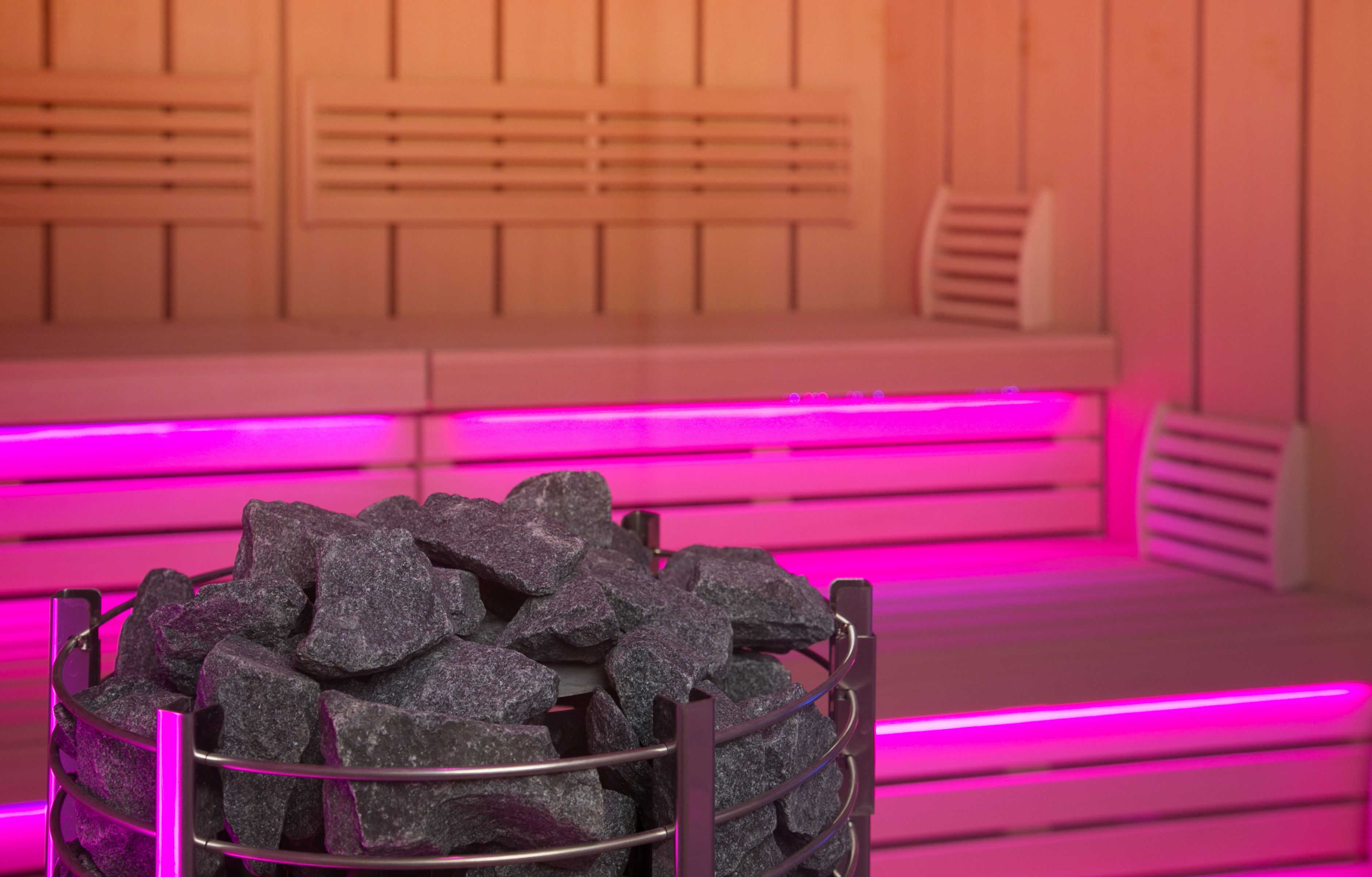 Uma sauna com suporte de metal cheio de pedras, bancos de madeira e iluminação rosa.