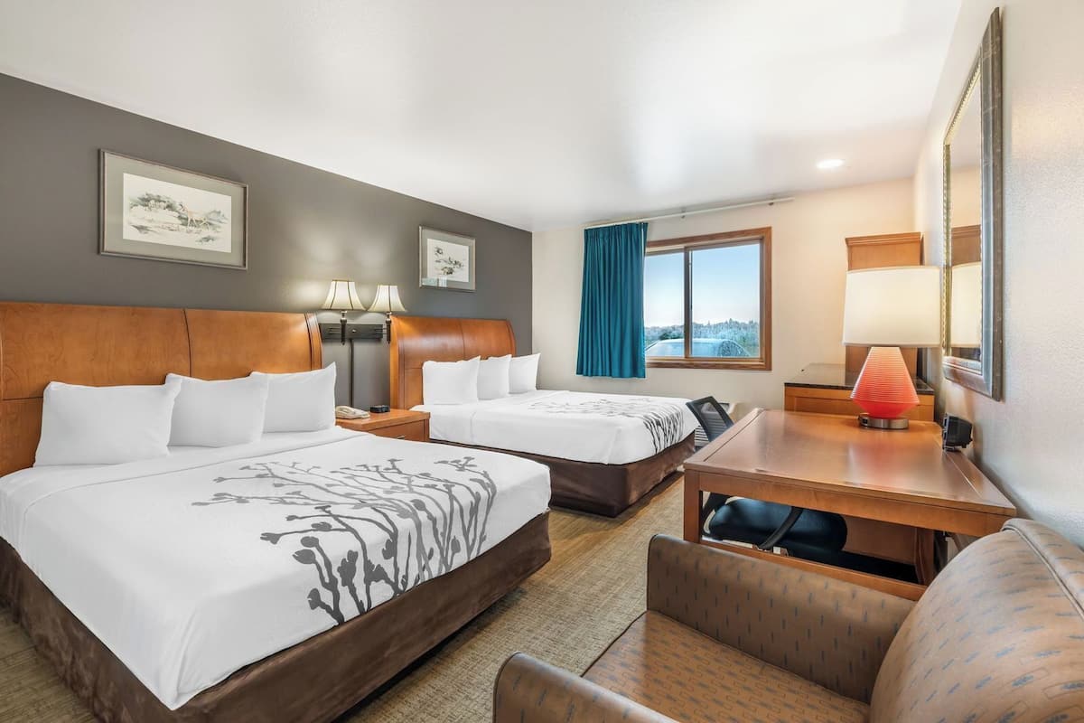 Quarto de hotel com duas camas, uma escrivaninha, uma cadeira, um abajur e uma janela grande.